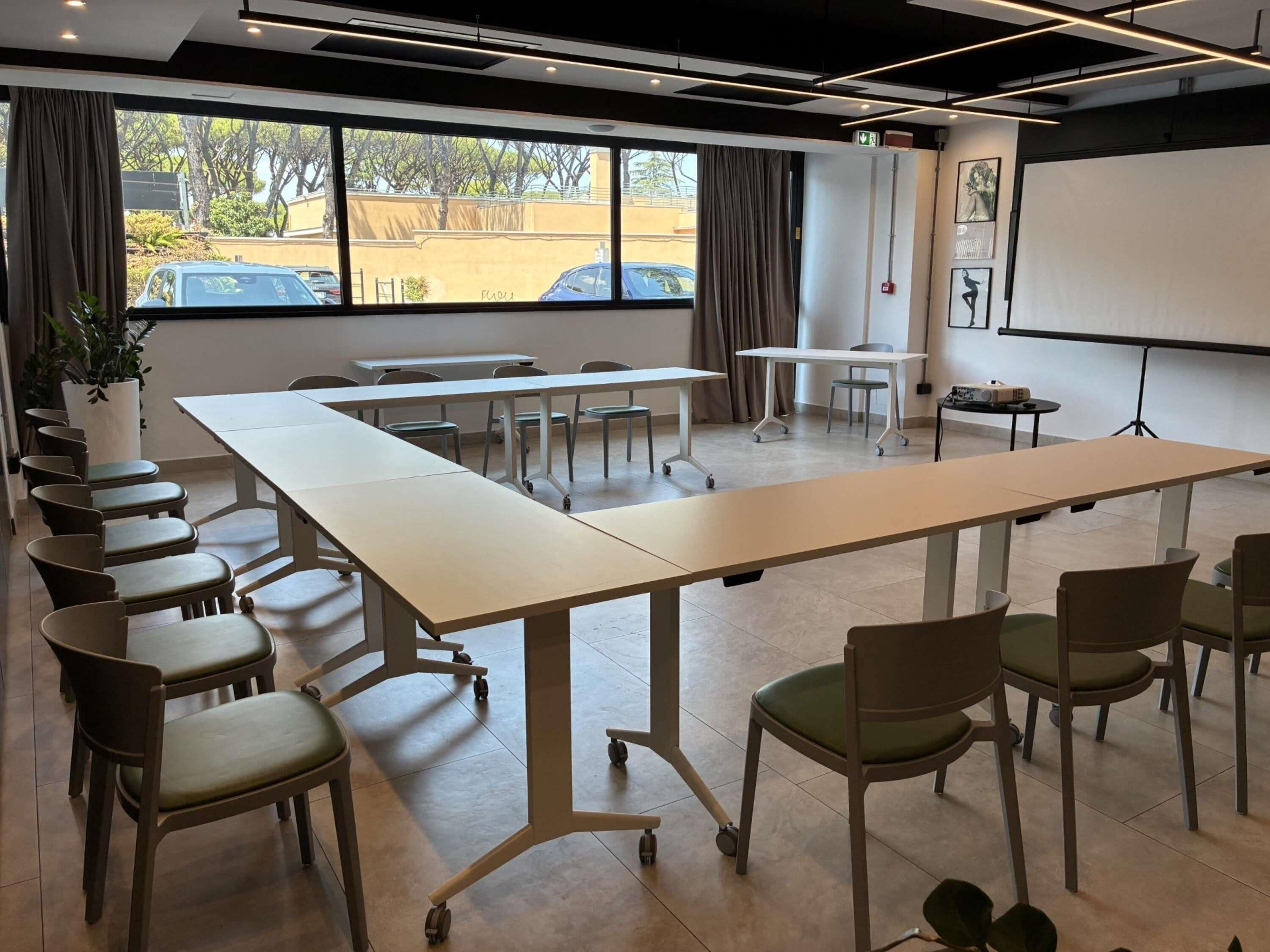 Uma sala de conferências com uma mesa comprida, cadeiras, um projetor e uma grande janela com vista para carros estacionados.