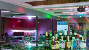 Uma área de bar com diversas garrafas de bebidas, um palco com equipamentos de música e um espaço de estar com cadeiras e mesas.