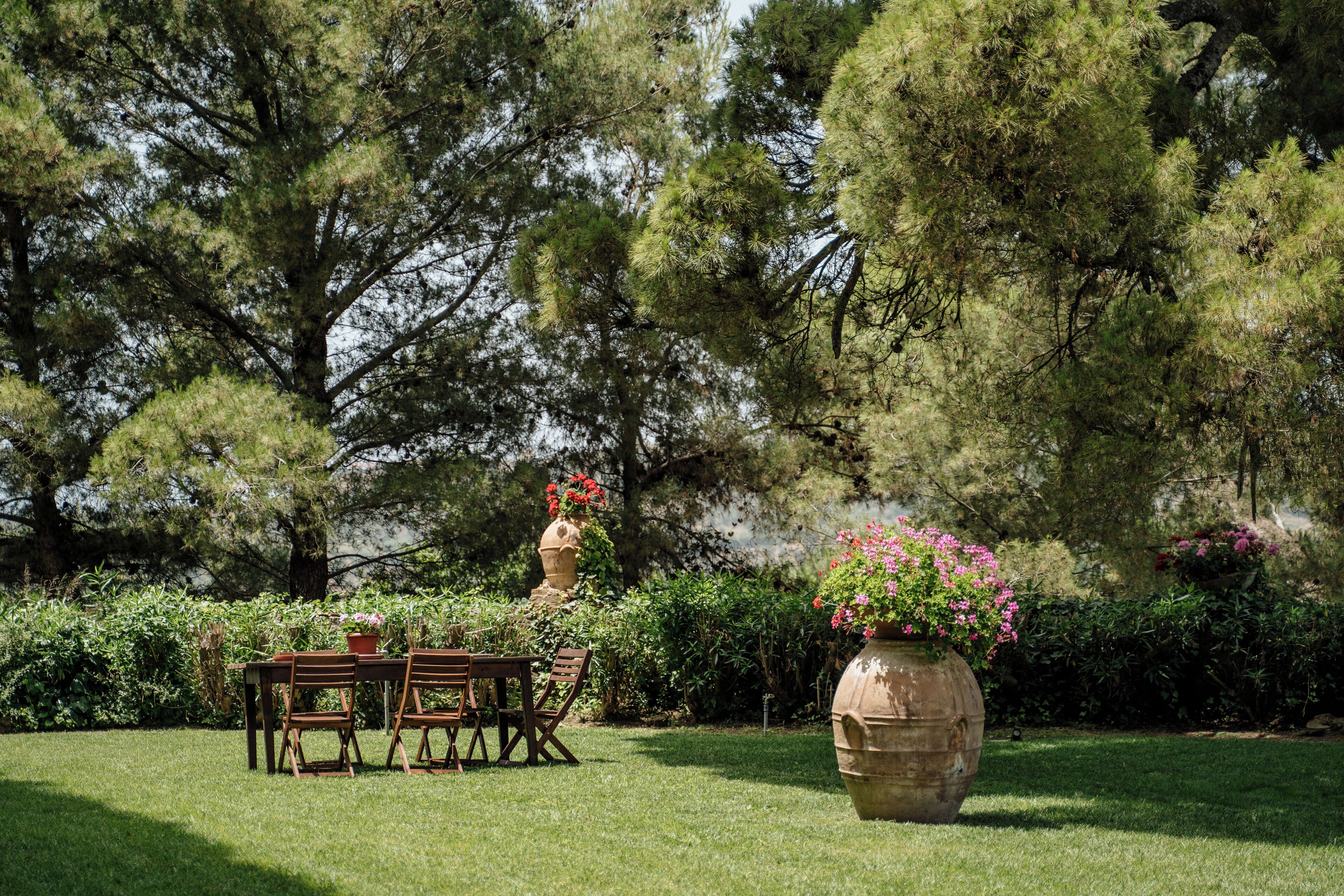 Um jardim com mesa e cadeiras de madeira, cercado por vegetação exuberante e um vaso grande de flores.
