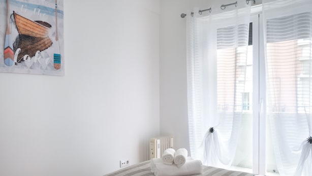 Quarto de hotel com cama, janela com cortinas finas, radiador e obra de arte na parede.