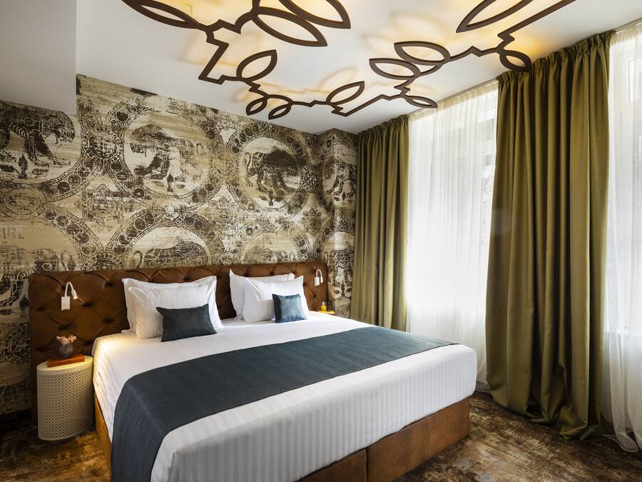 Quarto de hotel com uma cama grande, cabeceira com padrão decorativo, parede com design vintage e janela com cortinas.