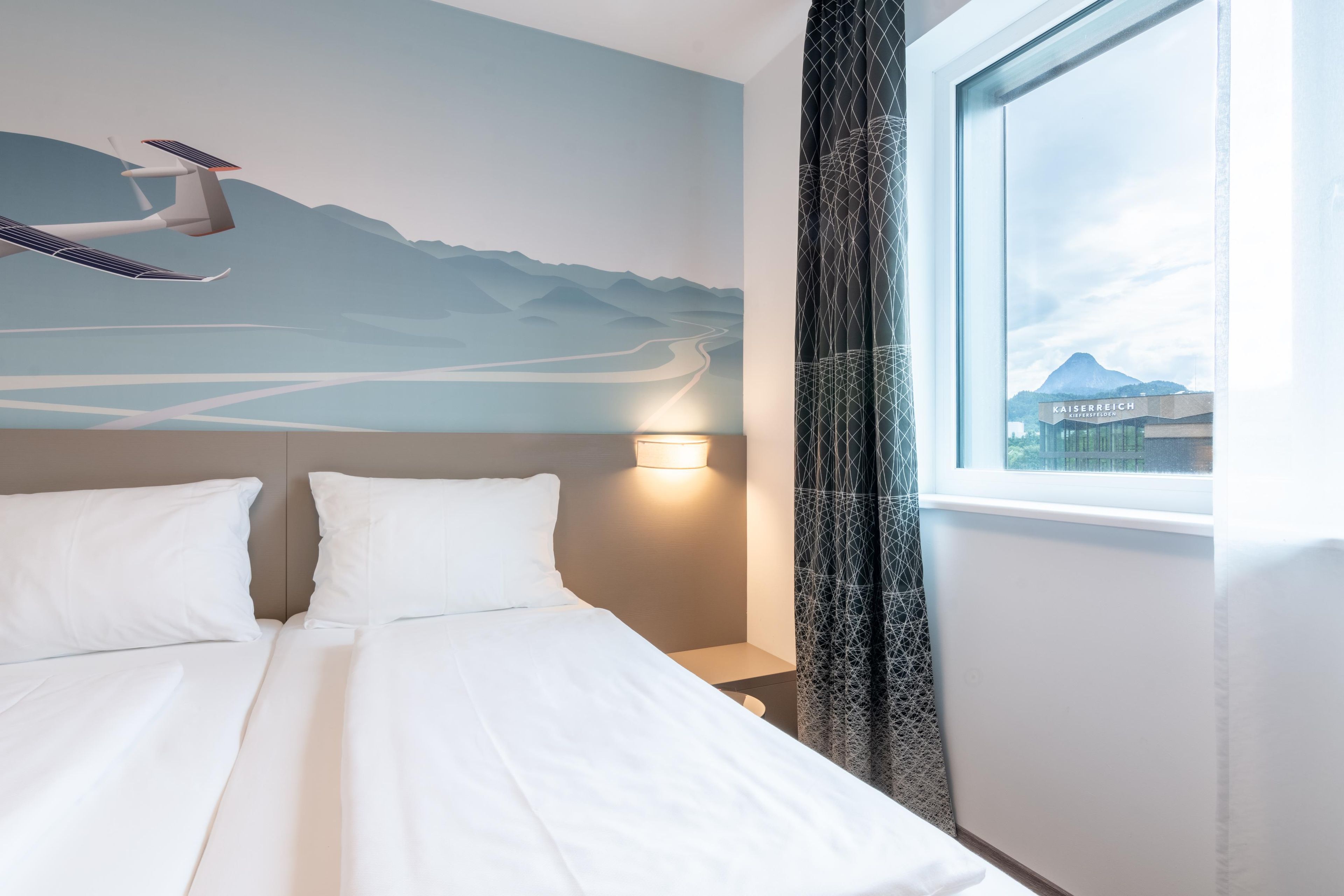 Quarto de hotel com cama, mesa de cabeceira, luminária na parede, janela com cortinas e um mural de paisagem montanhosa.