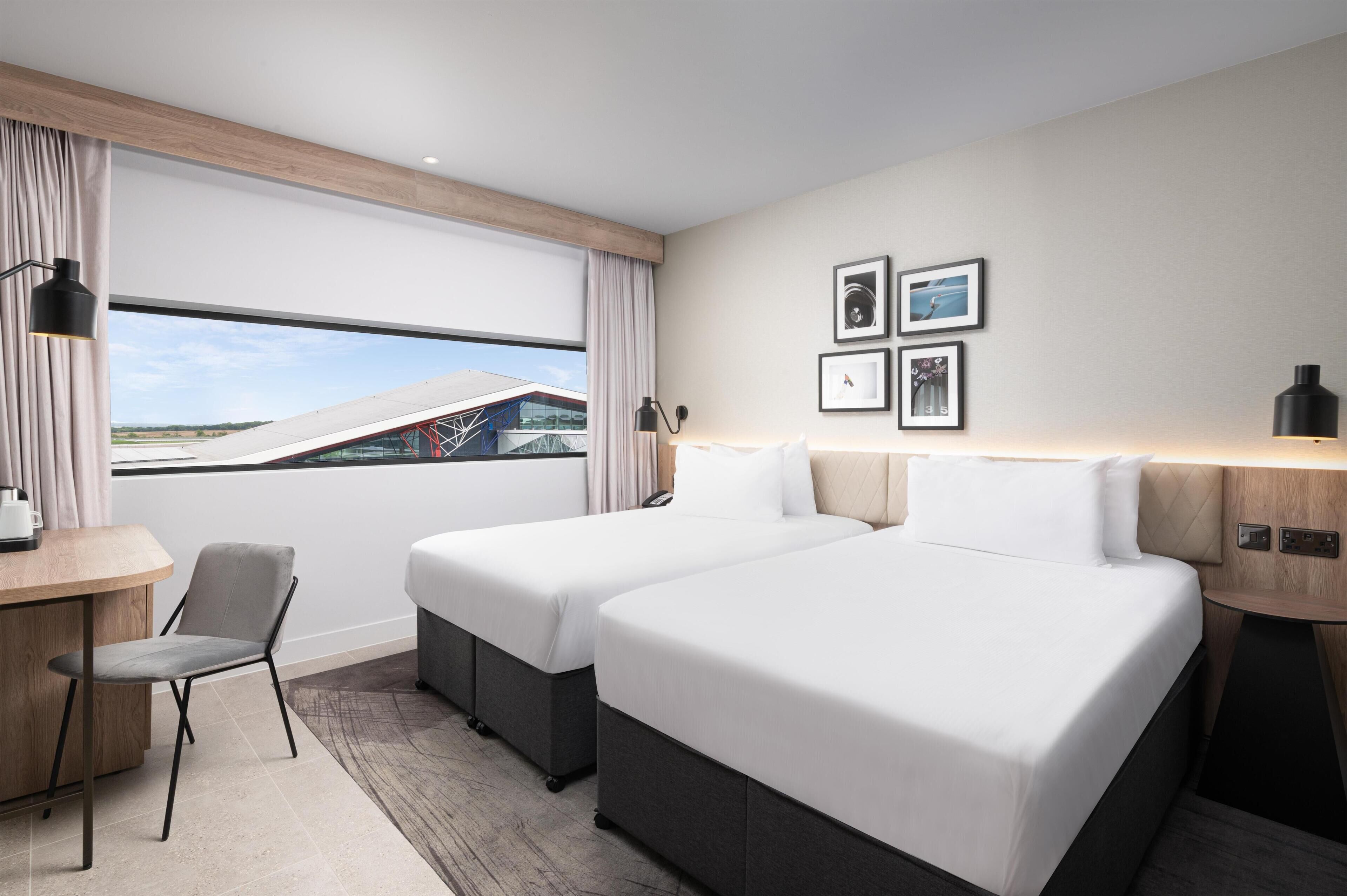 Quarto de hotel com duas camas, uma escrivaninha, uma cadeira, janela com cortinas e quadros nas paredes.