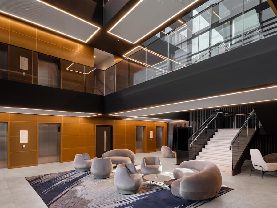 Um lobby moderno com área de estar central, escadaria e um mezanino com guarda-corpos de vidro.