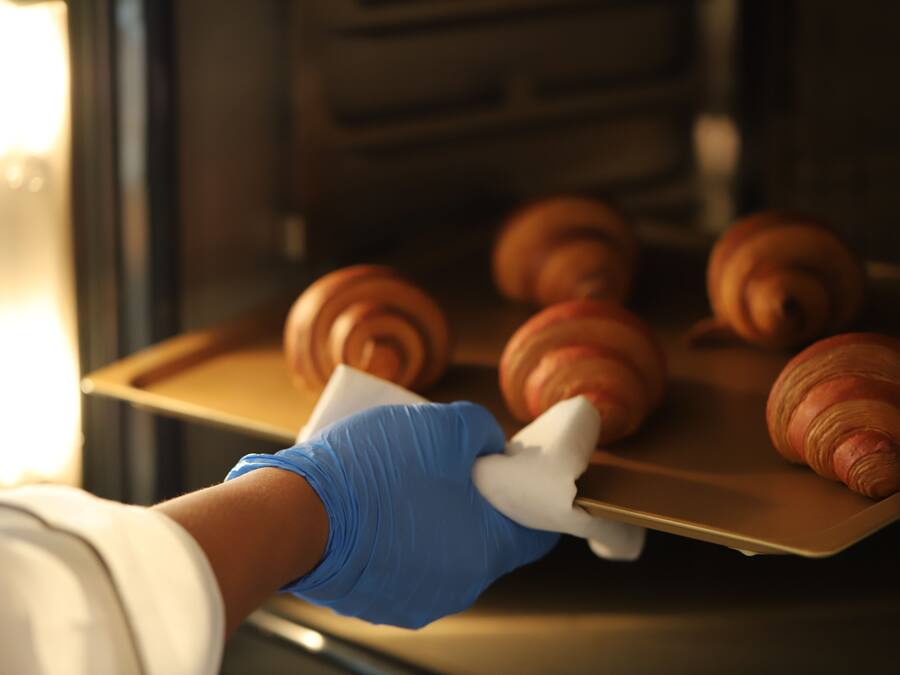 Pessoa usando uma luva azul manuseando uma bandeja com croissants dentro de um forno.
