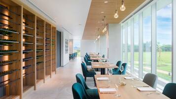 Uma sala de jantar moderna com mesas e cadeiras compridas, uma adega à esquerda e grandes janelas com vista para o exterior.