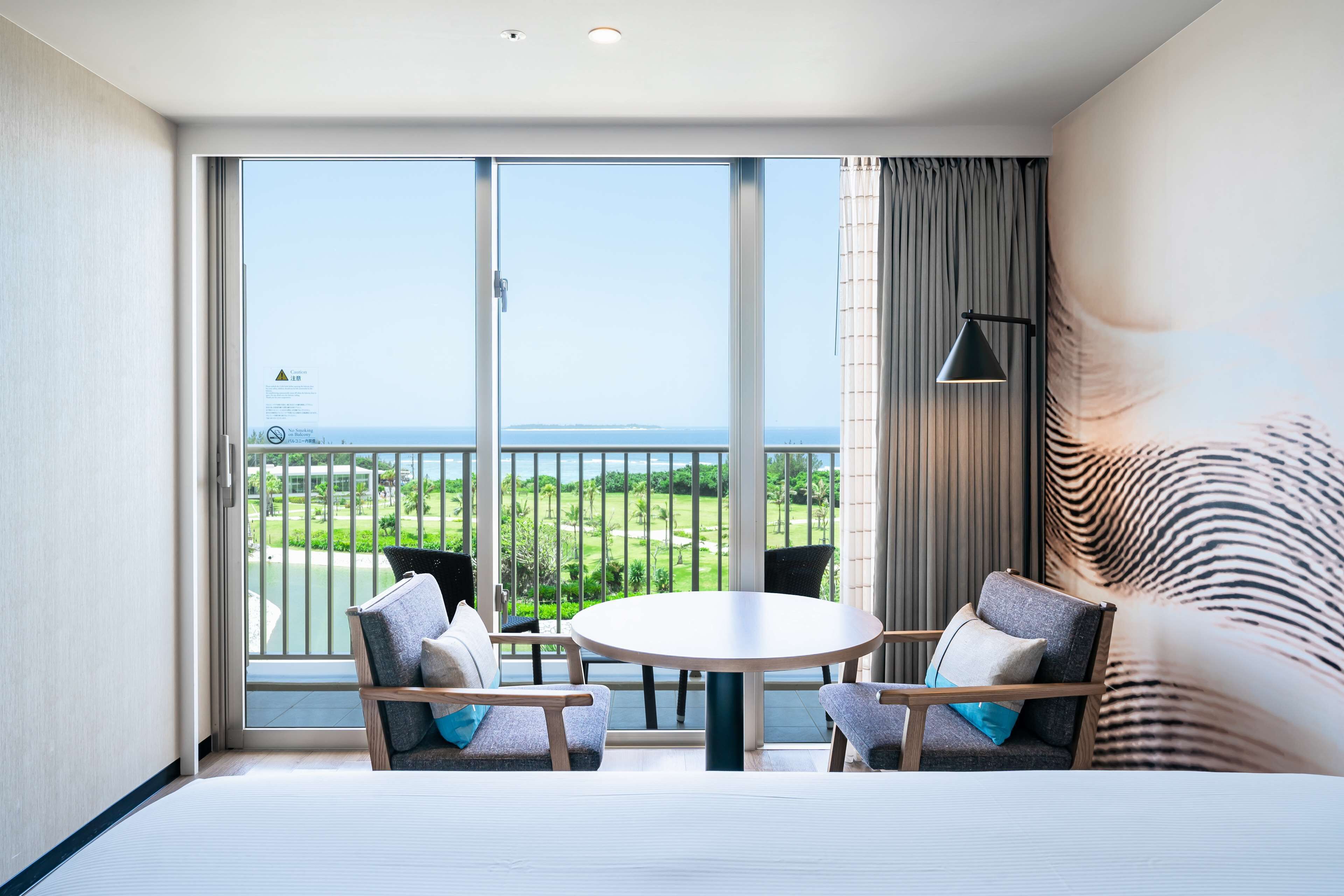 Quarto de hotel com uma grande porta de correr de vidro que dá para uma varanda com vista para o mar e vegetação. Há uma mesa redonda com duas cadeiras e um sofá com uma manta estampada.