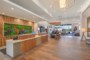 Uma área de recepção moderna com balcão de madeira, parede verde com plantas coloridas e um espaço de estar com vista para o exterior.