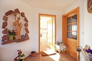 Um quarto com porta de madeira, uma pequena mesa com toalha, um vaso de planta e uma estátua em uma prateleira.