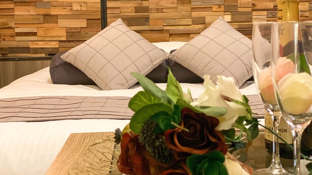 Uma cama bem arrumada com duas almofadas, um vaso com flores e duas taças de vinho sobre uma mesa.