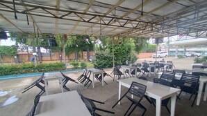 Um café ao ar livre com mesas e cadeiras, um telhado coberto e vista para a rua e vegetação.