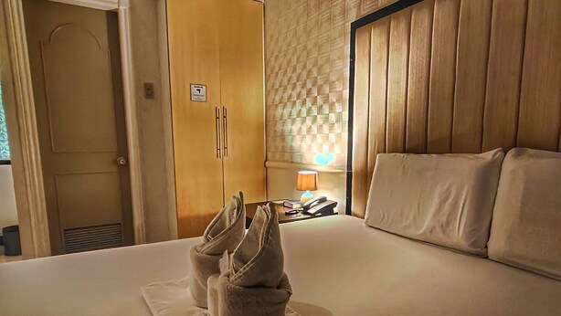 Quarto de hotel com uma cama grande, um guarda-roupa de madeira, uma mesa de cabeceira com luminária e uma porta com placa.