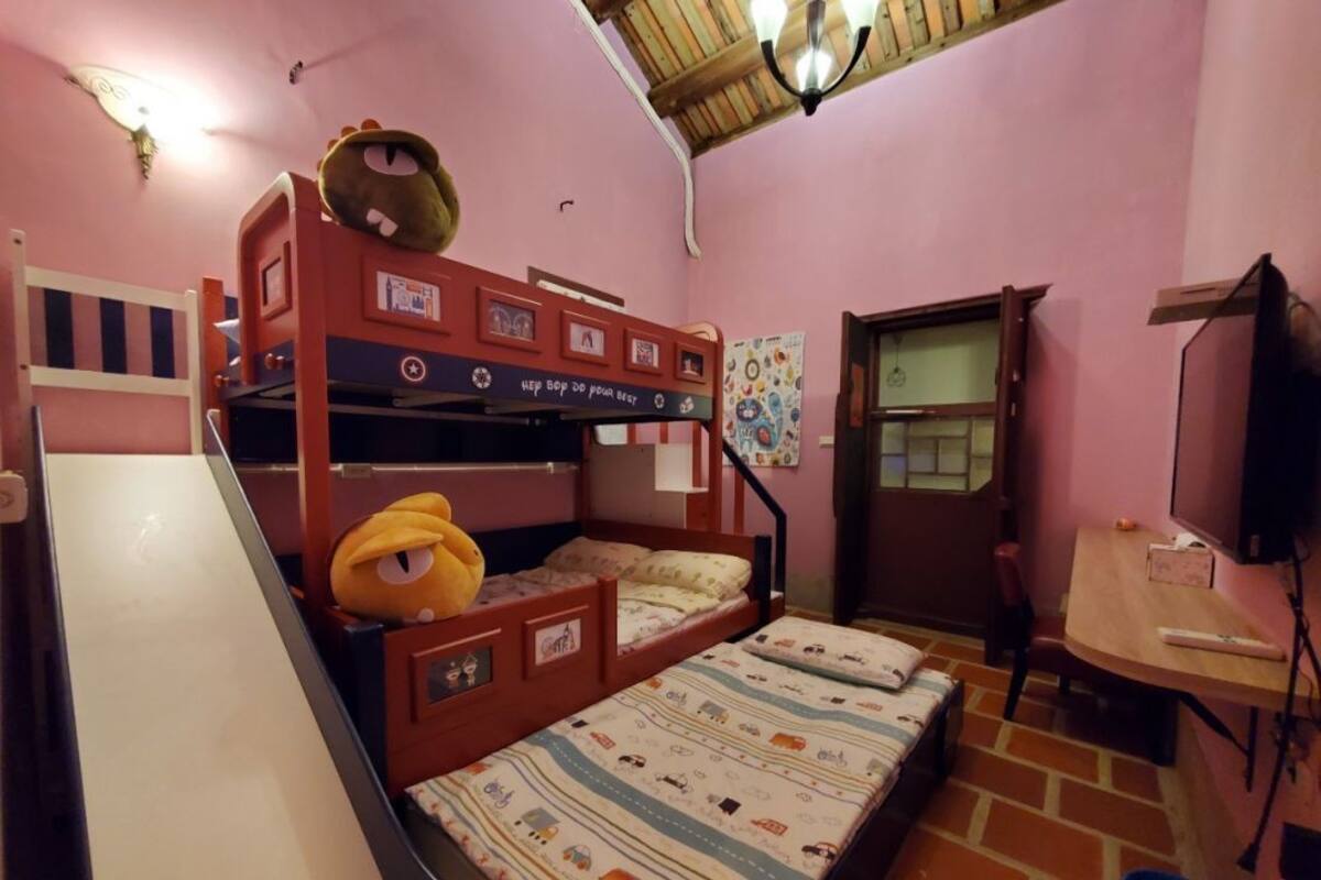 Quarto com beliche, escrivaninha e cadeira, uma janela pequena e ventoinha no teto.
