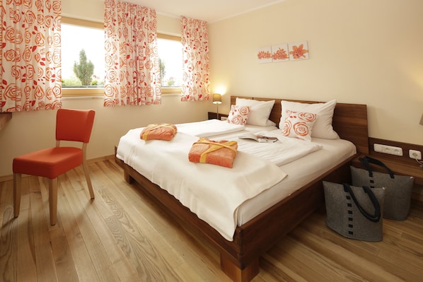 Cama bem arrumada com colcha branca e laranja estampada, cabeceira de madeira e duas almofadas com padrões combinando. Há uma cadeira vermelha à esquerda da cama, uma mesa de cabeceira com abajur e uma janela com cortinas estampadas.