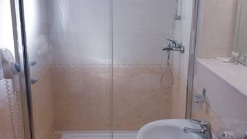 Um chuveiro com divisória de vidro, um chuveiro manual, uma pia com espelho e uma pequena janela na parte superior da porta do chuveiro.