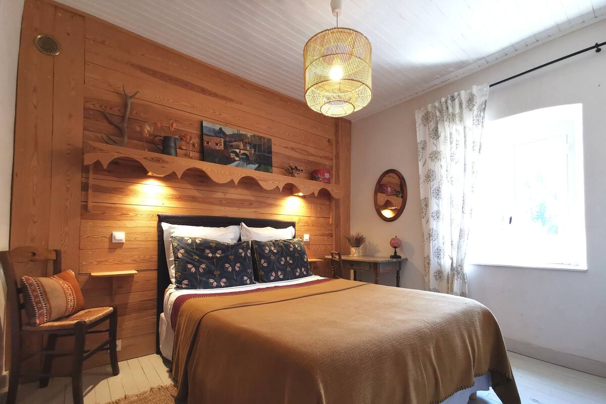 Um quarto aconchegante com cabeceira de madeira, uma cama grande com manta marrom, uma luminária pendente de vime e uma janela com cortinas.
