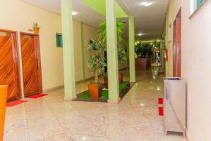 Corredor de hotel com portas de madeira, pilares verdes, plantas em vasos e um extintor de incêndio.