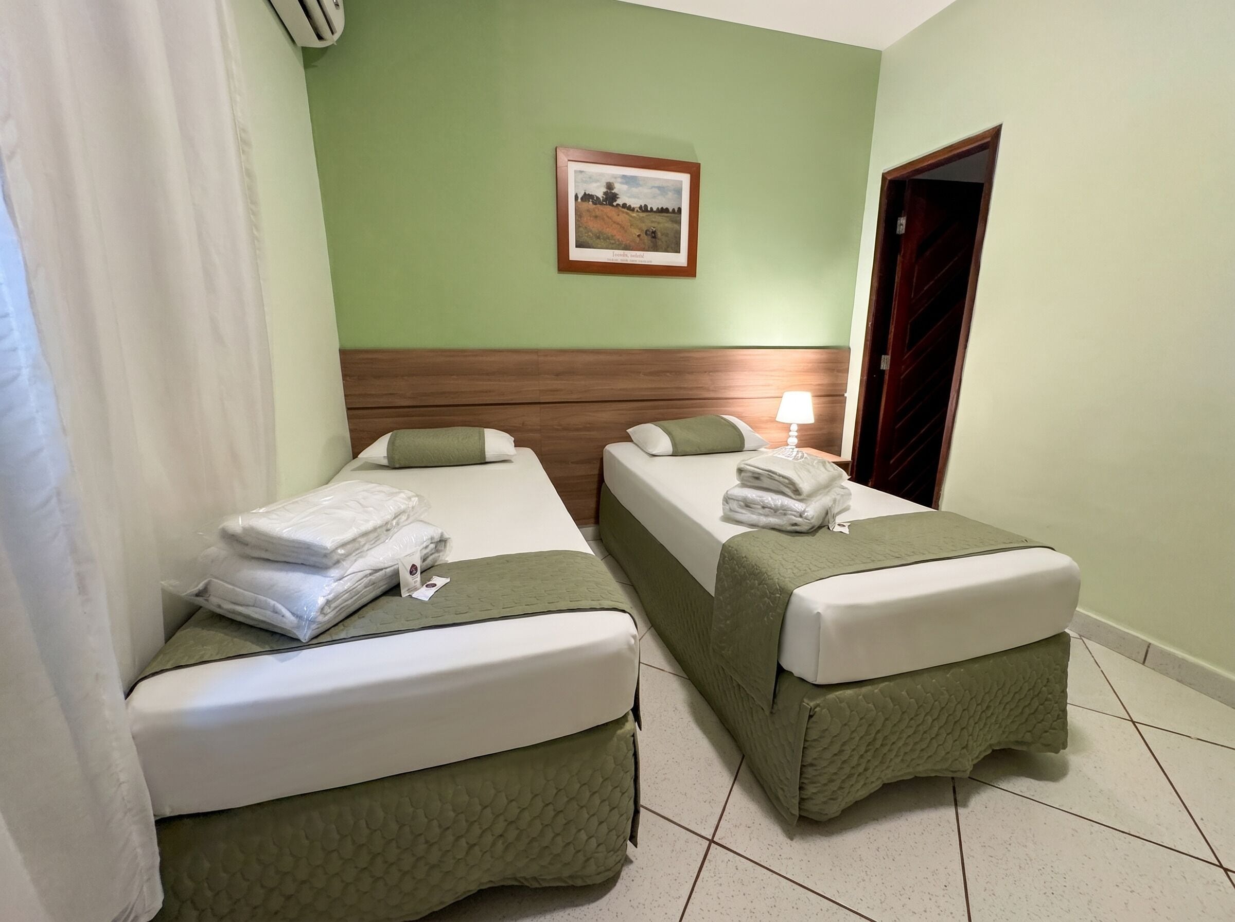 Quarto de hotel com duas camas, um quadro na parede e uma porta à direita.