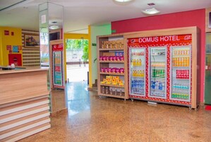 Uma área de recepção de hotel com balcão, placa Domus Hotel e máquinas de venda automática de bebidas.