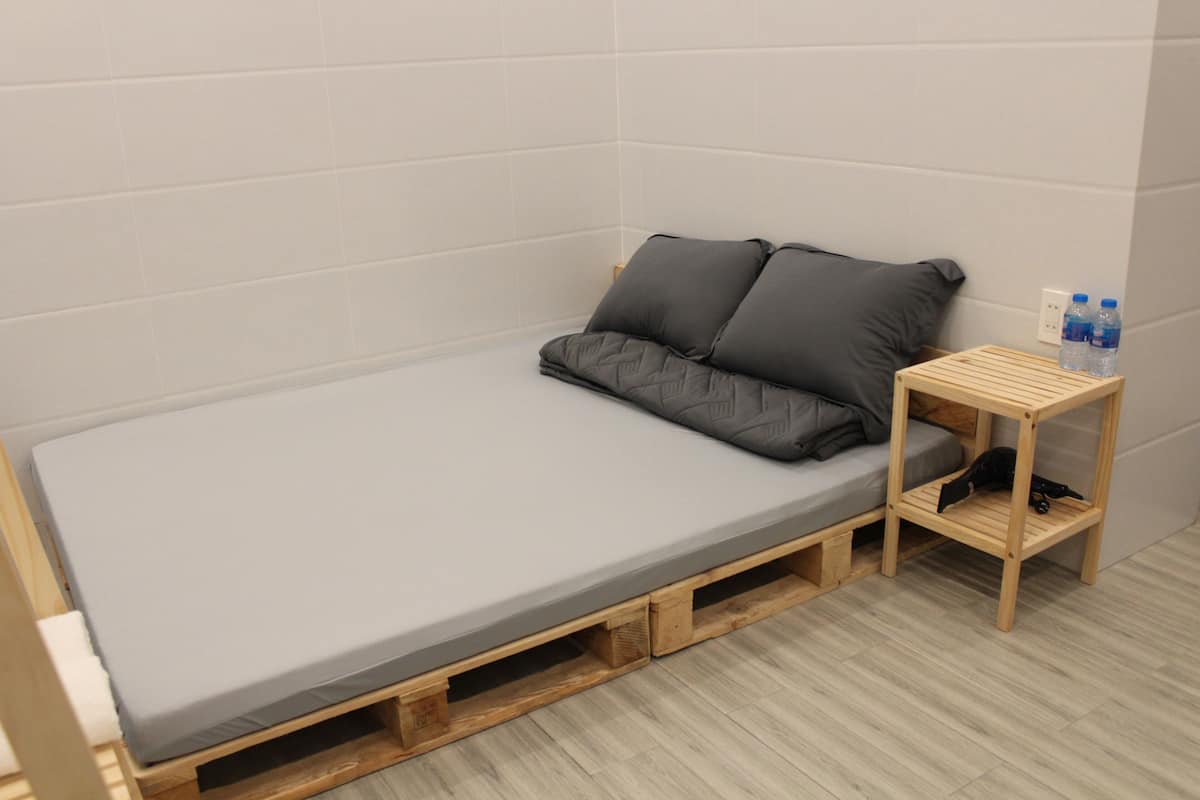 Quarto pequeno com cama de solteiro, colchão e duas almofadas cinzas, além de mesa de cabeceira de madeira com duas garrafas de água.