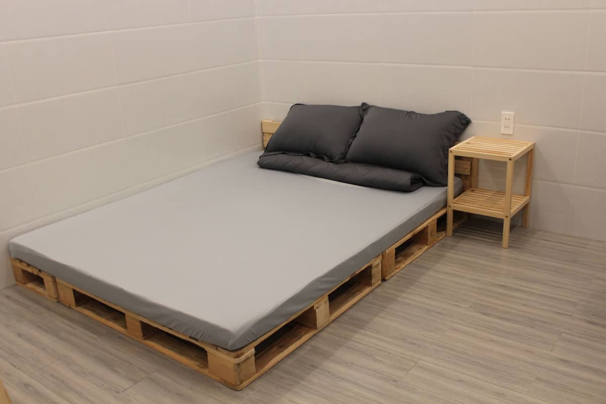 Quarto pequeno com uma cama de solteiro, estrutura de madeira, colchão e duas almofadas cinzas, além de uma pequena mesa de cabeceira de madeira.