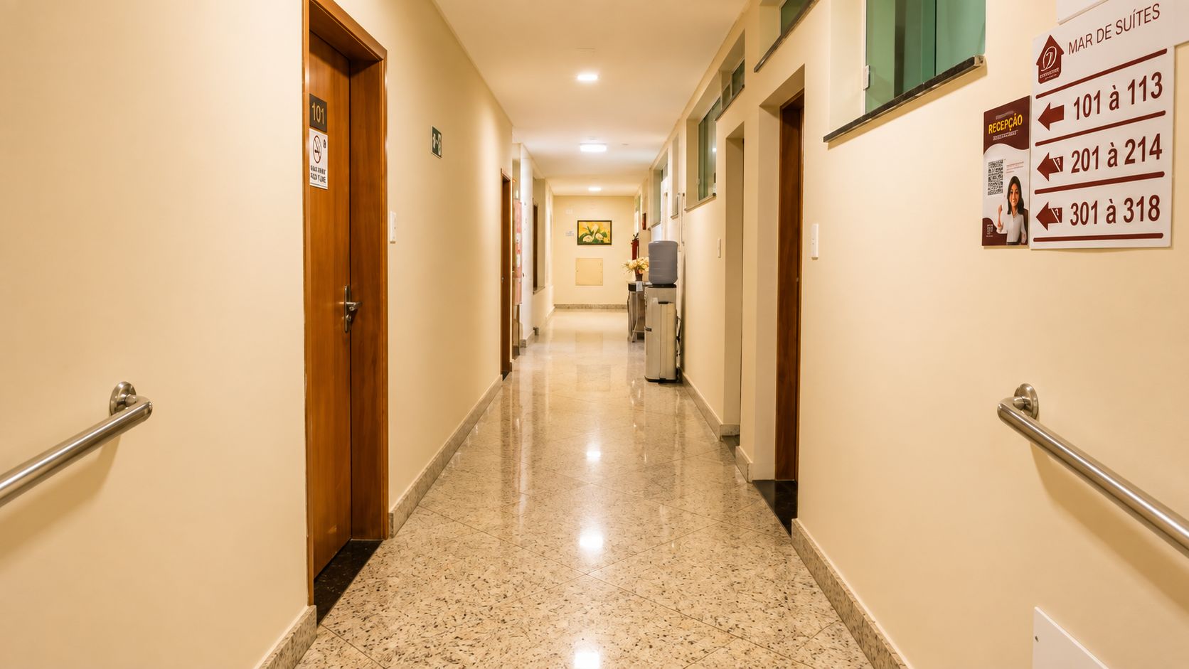 Corredor de hotel com portas numeradas de 101 a 318, corrimãos em ambos os lados e placa de recepção.