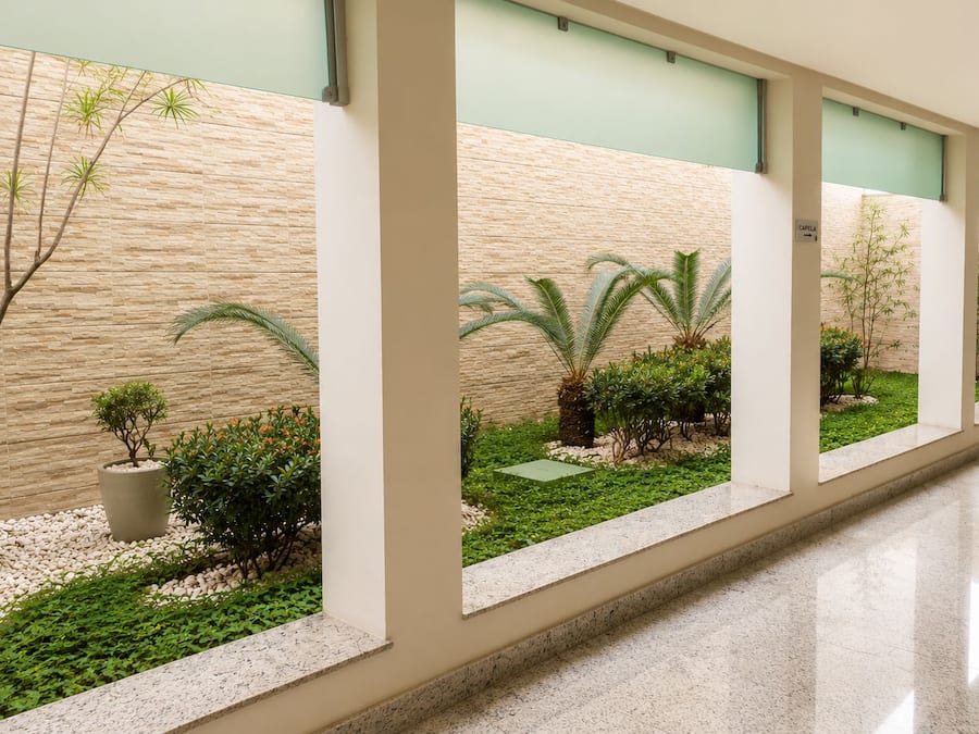 Um corredor com várias janelas, cada uma oferecendo vista de um jardim ajardinado com diversas plantas e chão coberto de pedrinhas.