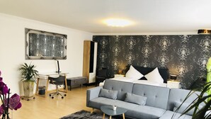 Quarto moderno com sofá cinzento, cama com roupa de cama branca, escrivaninha com cadeira e uma grande obra de arte emoldurada na parede.