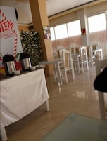 Uma área de refeições com uma mesa posta para uma refeição, cercada por cadeiras brancas. Há uma faixa com texto e uma planta em vaso ao fundo.