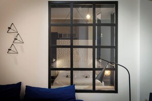 Quarto com uma janela grande, uma cama, uma escrivaninha e um abajur.
