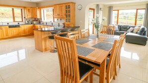 Uma cozinha espaçosa com armários de madeira e uma ilha central, uma sala de jantar com uma mesa grande e cadeiras, e uma sala de estar com sofá e vista para o exterior.