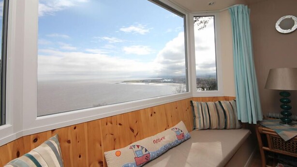 Quarto com uma janela ampla com vista para um corpo de água, um banco de madeira com almofadas e uma luminária sobre uma mesinha.