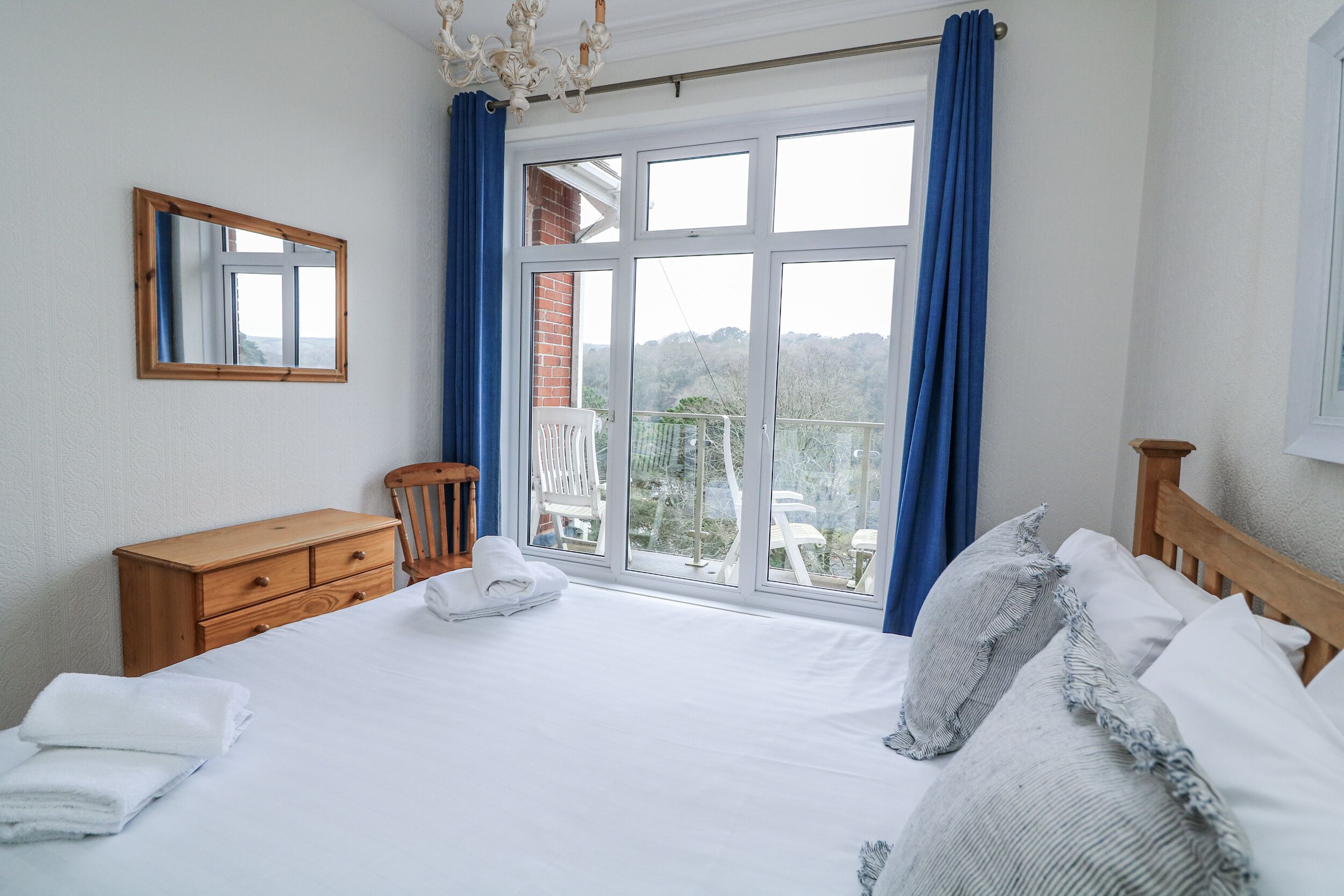 Quarto com cama grande, cómoda de madeira, uma cadeira e varanda com cortinas azuis.