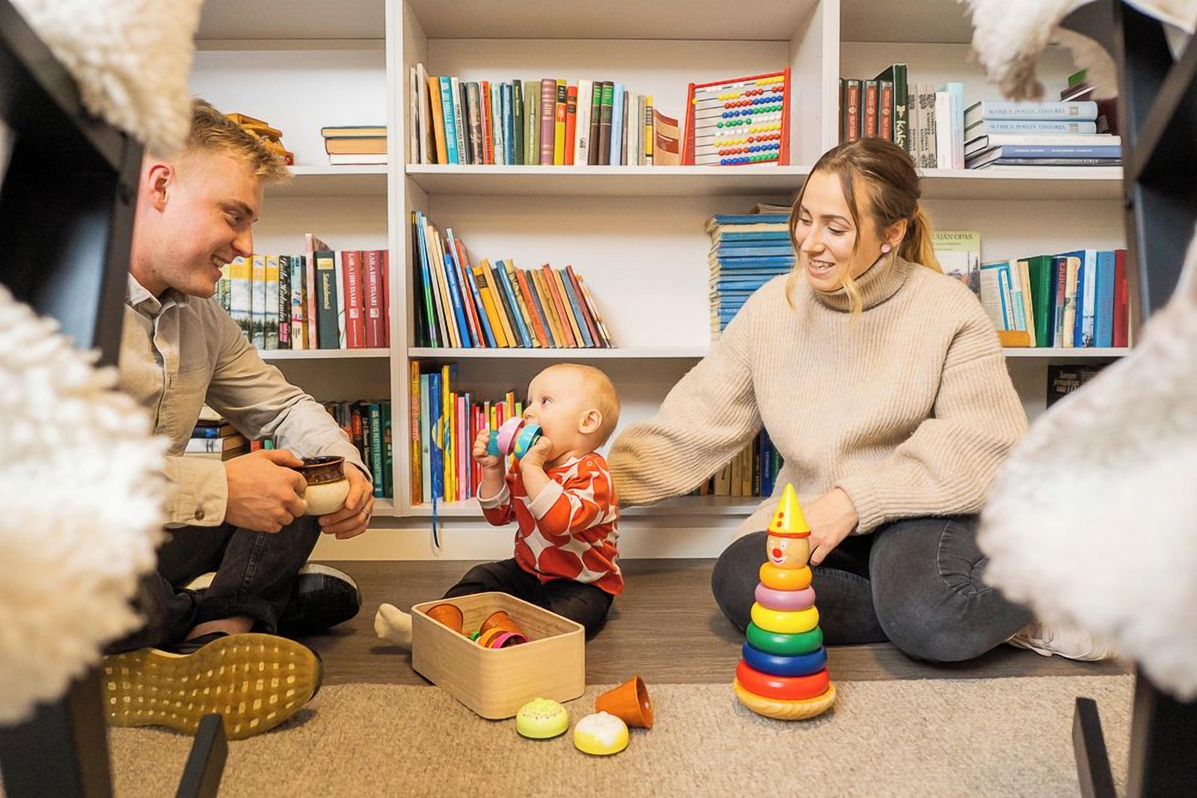 Quarto com estante cheia de livros, um bebê com brinquedos e uma pessoa sentada no chão.