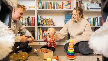 Quarto com estante cheia de livros, um bebê com brinquedos e uma pessoa sentada no chão.