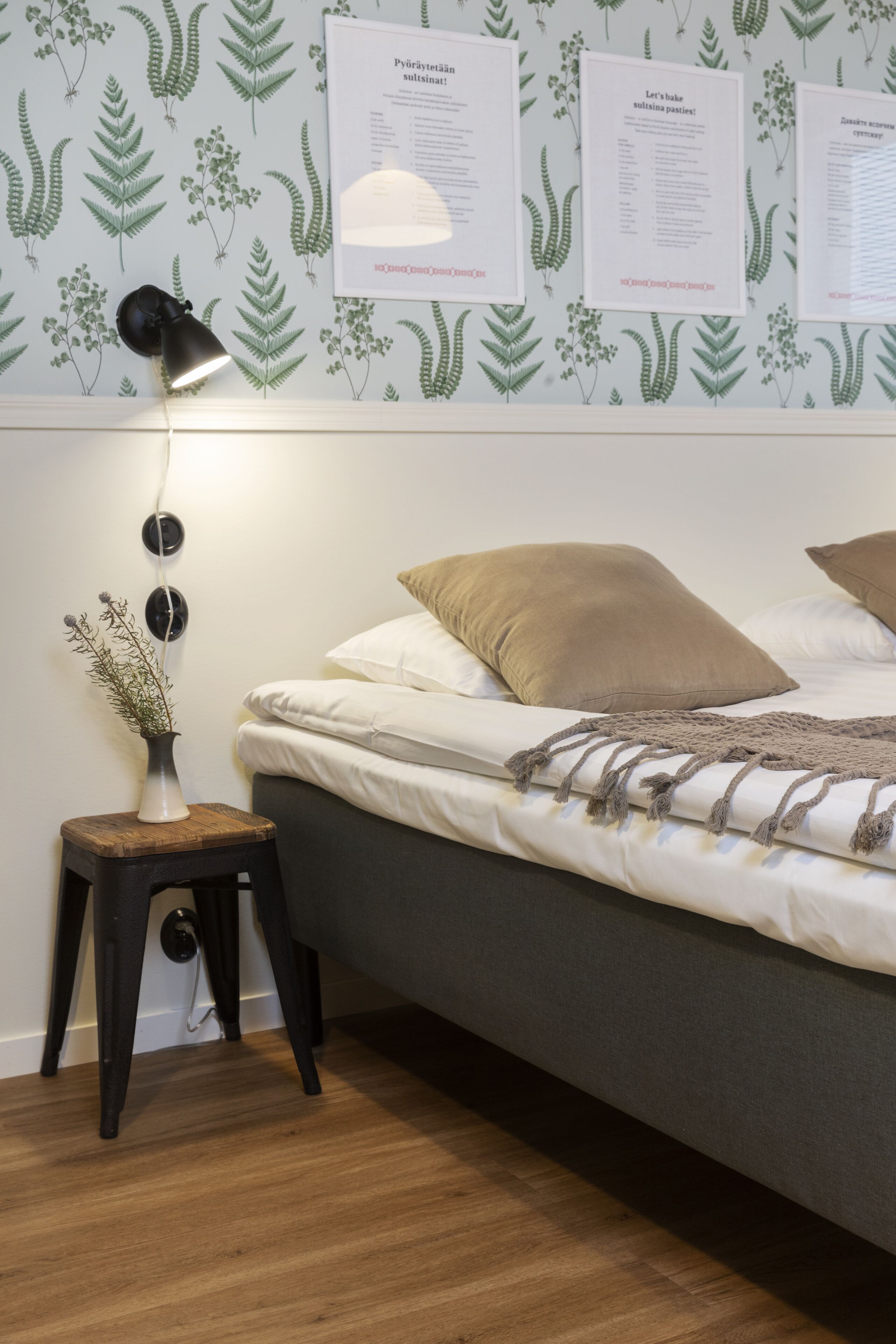 Cama bem arrumada com cabeceira cinza, abajur e pequeno banquinho de madeira com um vaso de flores secas.
