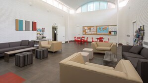 Um lobby espaçoso com teto alto, paredes de tijolo branco e uma grande janela que proporciona luz natural. Há várias áreas de estar com sofás e pufes, e um mural de avisos com mapas e comunicados.