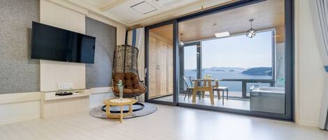 Uma sala de estar moderna com televisão de tela plana, uma poltrona de couro marrom, uma mesa de madeira redonda e vista para o mar através de grandes portas de vidro.