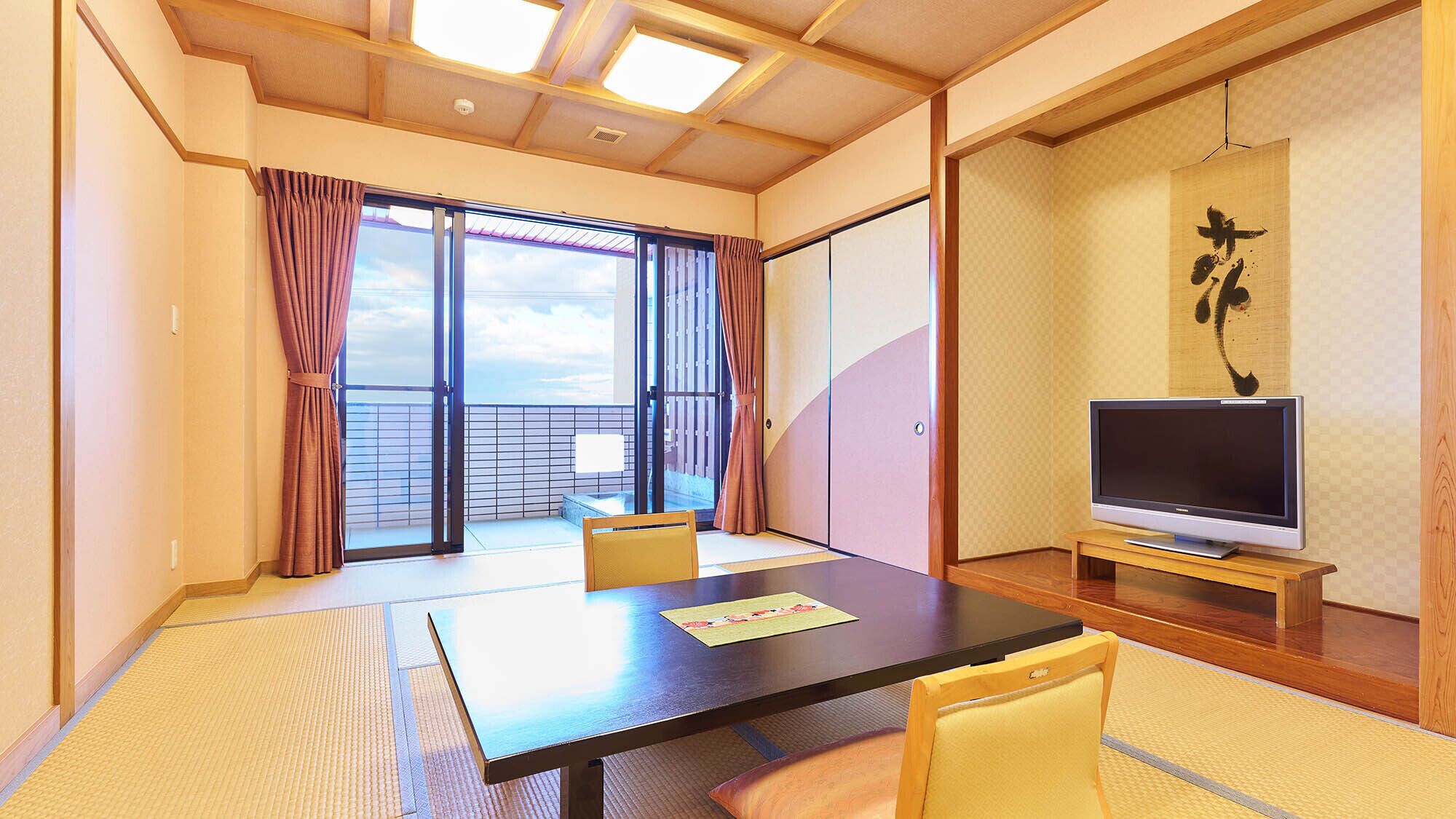 Quarto com uma mesa grande, duas cadeiras, uma TV e uma porta de correr para uma varanda.