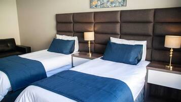 Quarto de hotel com duas camas, cada uma com colcha azul e lençóis brancos. Há duas mesas de cabeceira com luminárias.