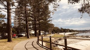 Uma passarela pavimentada ao lado de uma praia, com guarda-corpo de madeira, um carro estacionado e um edifício ao fundo.