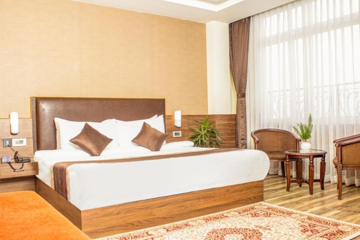 Quarto de hotel com cama de cabeceira em madeira, mesa de cabeceira com luminária, pequena mesa redonda com uma planta, uma cadeira e uma janela grande com cortinas.