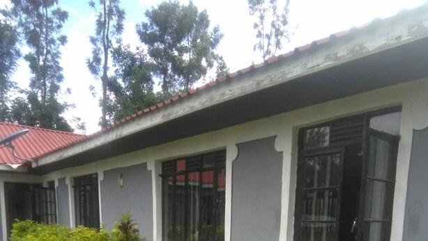 Um edifício de dois andares com fachada cinza e telhado vermelho. Possui duas portas visíveis, uma no andar térreo e outra no andar superior. O edifício tem um pequeno jardim frontal com arbustos verdes e um caminho de concreto que leva à entrada.