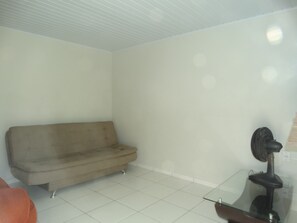 Quarto com sofá bege, mesa de vidro e um ventilador.