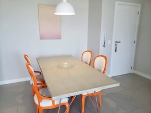 Uma mesa de jantar com quatro cadeiras laranjas, uma luminária pendente branca e uma porta com fechadura.