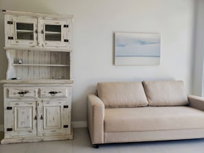 Um armário de madeira branca com aparência desgastada, com várias gavetas e prateleiras, um sofá bege e um quadro com uma paisagem na parede.