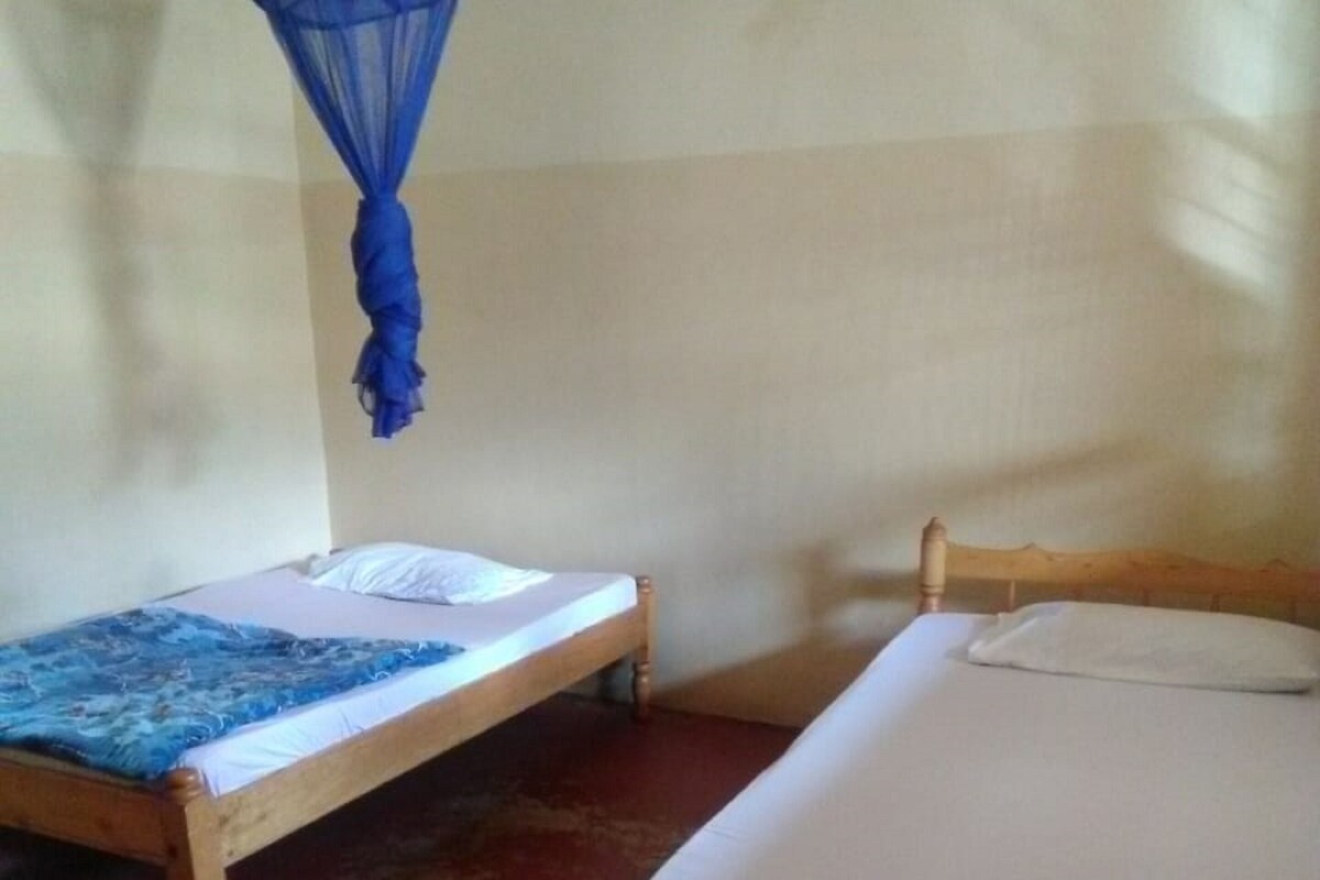 Quarto com duas camas de madeira, uma com colcha azul e outra com colcha branca. Um par de chinelos vermelhos está no chão.