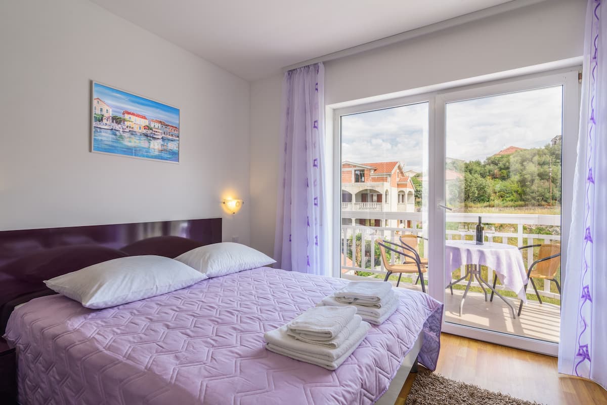 Quarto com cama, almofadas, um quadro, uma varanda com mesa e cadeiras, e vista para edifícios e vegetação.