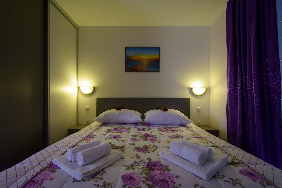 Quarto de hotel com uma cama, duas luminárias de cabeceira, um quadro na parede e uma cortina roxa.