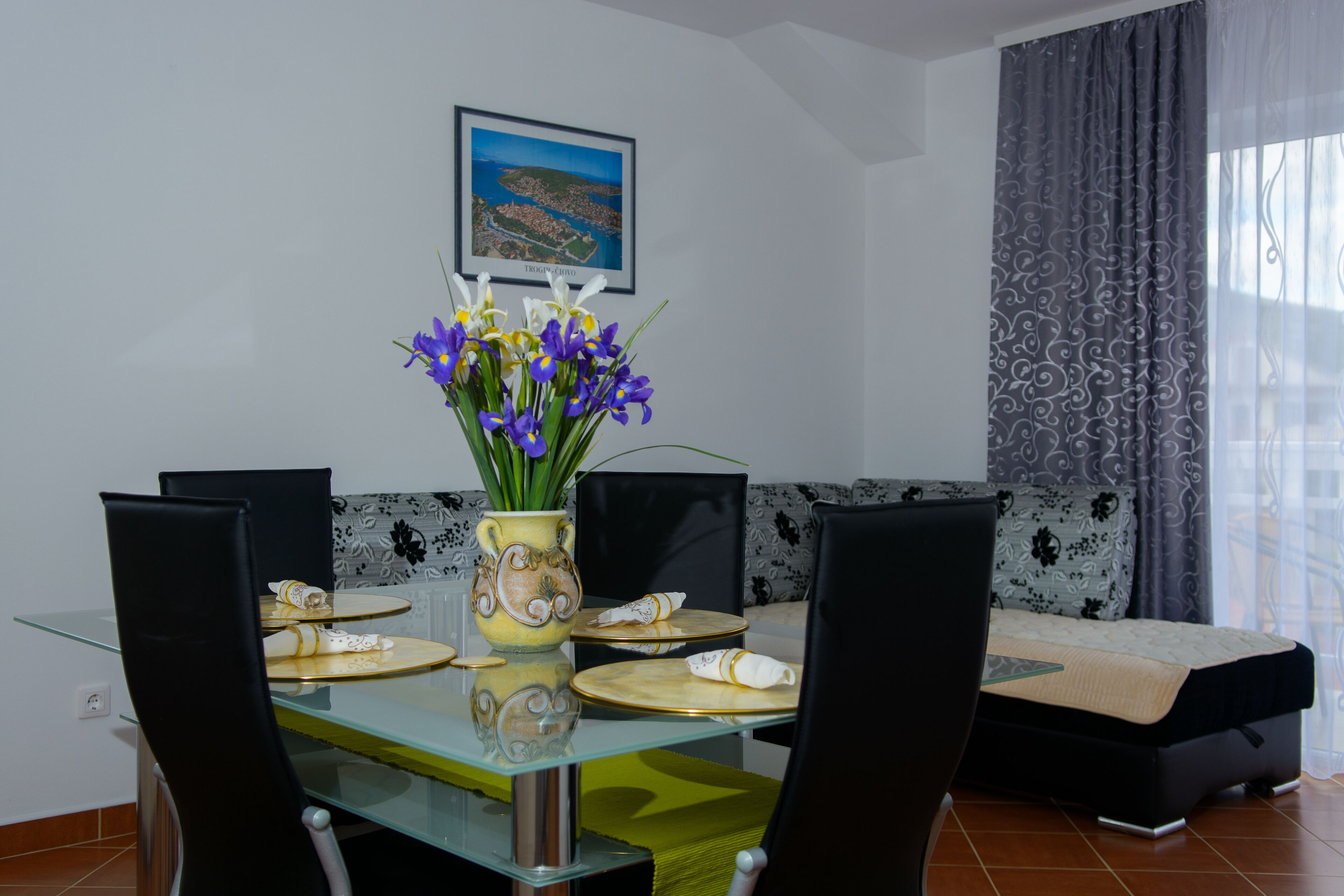 Uma sala de jantar com mesa de vidro, cadeiras e um vaso com flores. Há uma cama com colcha estampada e cortinas.